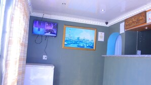 Quarto com televisão na parede, quadro com vista urbana, geladeira branca e bancada com pia.