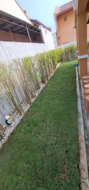 Uma estreita trilha gramada ladeada por bambus e uma parede branca, conduzindo a um edifício com telhado de azulejos.