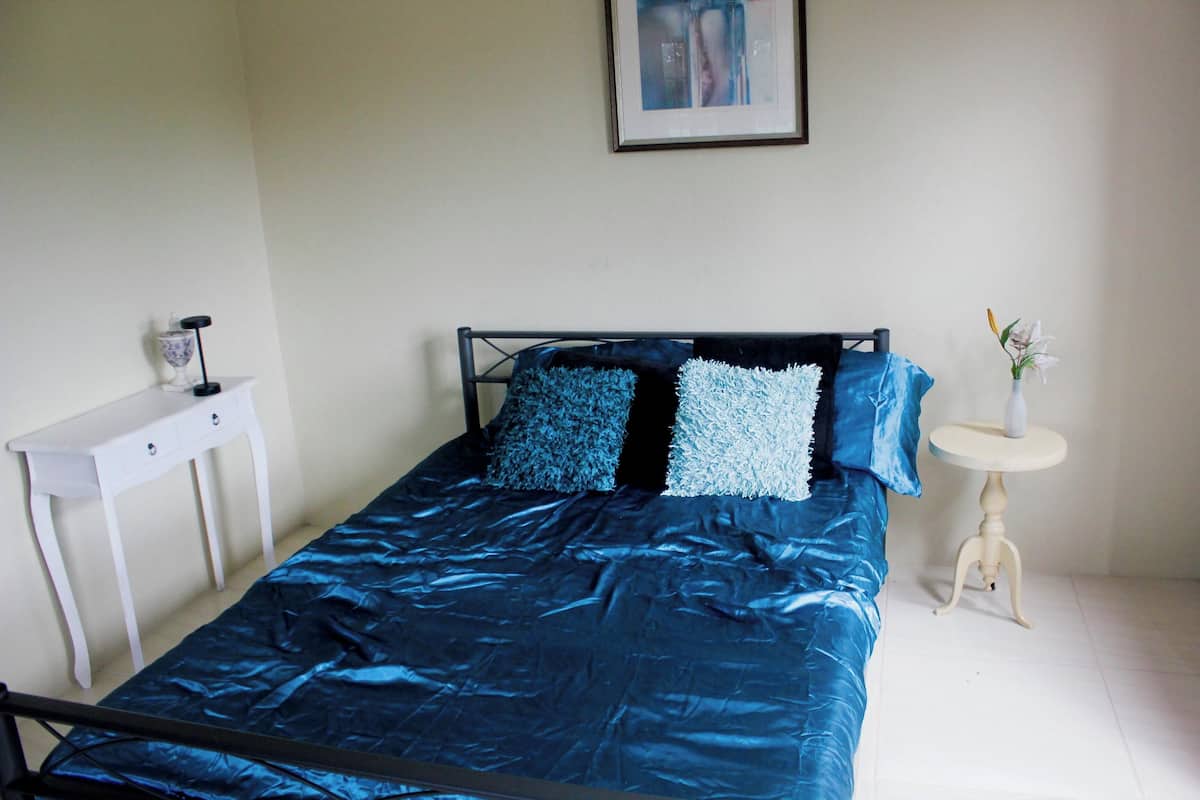 Um quarto com cama, mesa de cabeceira, uma mesinha com um vaso e um quadro na parede.