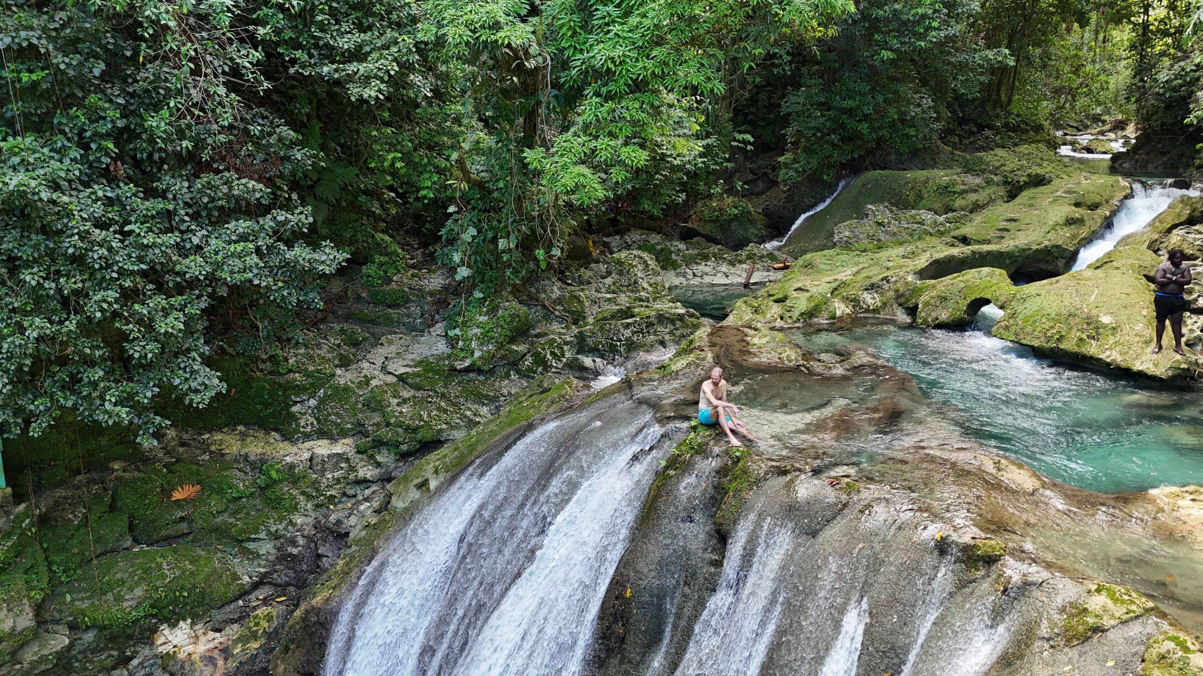 Uma cascata em uma área arborizada com uma pessoa sentada em uma rocha.