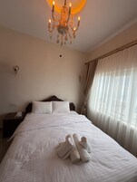 Quarto de hotel com uma cama grande, um candelabro, uma mesa de cabeceira e uma janela com cortinas.