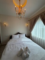 Quarto de hotel com uma cama grande, um lustre e cortinas numa janela.