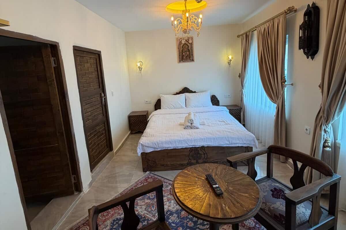 Quarto de hotel com cama grande, lustre, mesa redonda com cadeiras e tapete estampado.