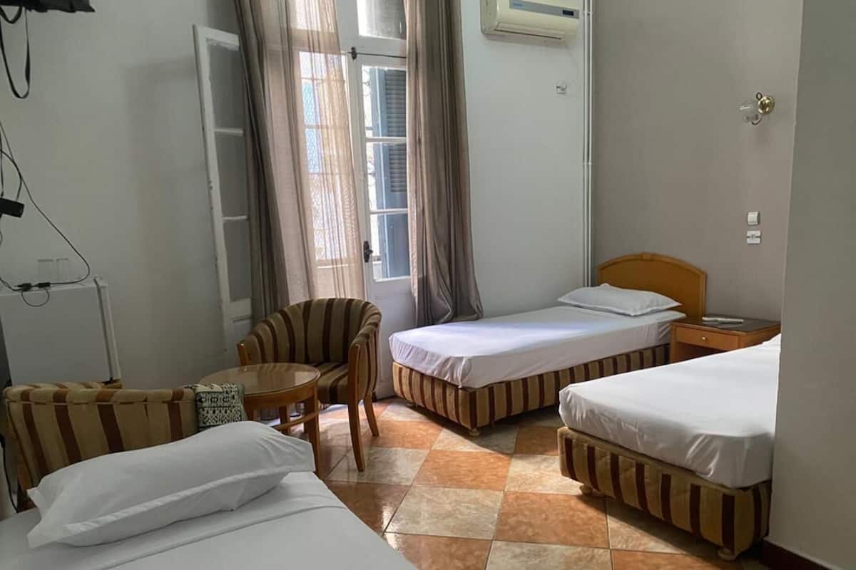 Quarto de hotel com duas camas, uma cadeira, uma mesinha e uma janela com cortinas.