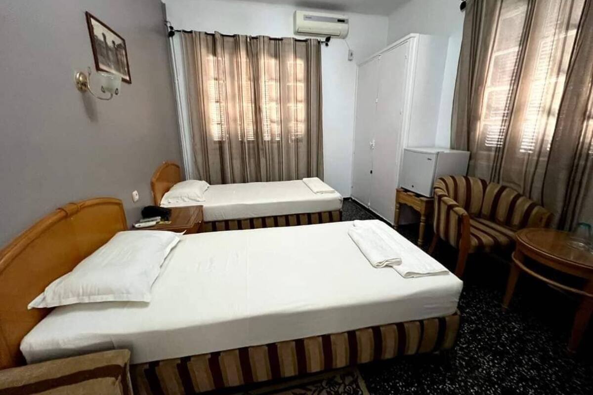 Quarto de hotel com duas camas, um sofá, uma mesa pequena, ar condicionado e uma janela com cortinas.