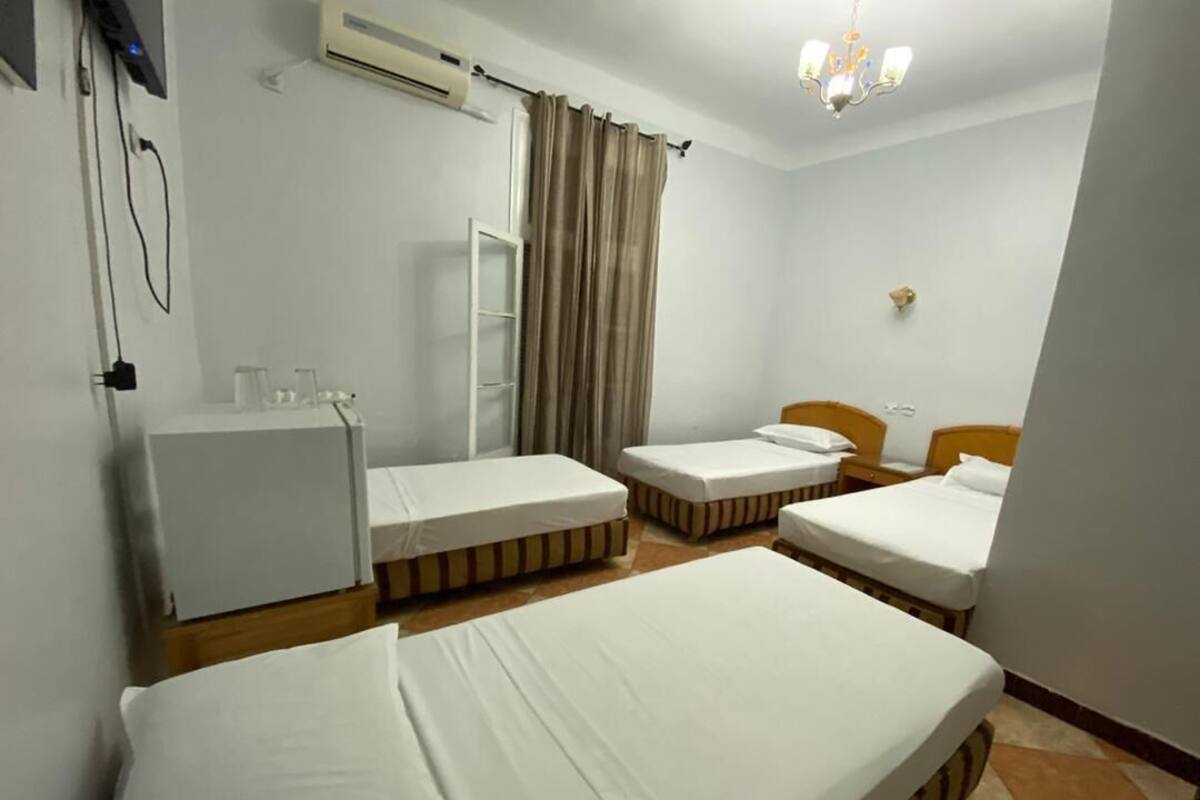 Quarto de hotel com três camas de solteiro, uma pequena mesa, uma cadeira, um guarda-roupa e um aparelho de ar condicionado.