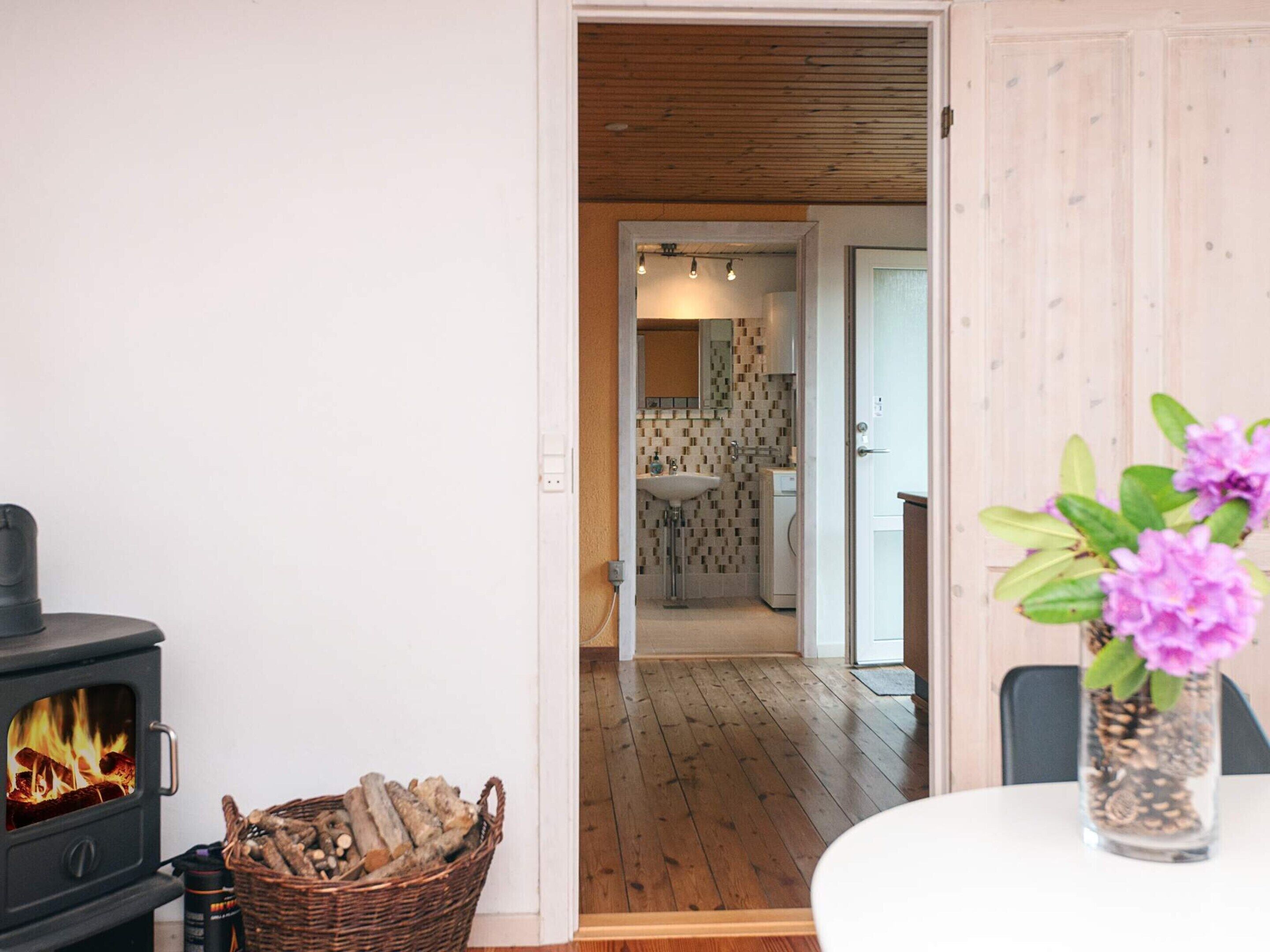 Um quarto com uma lareira a lenha, um cesto de vime com lenha, uma mesa branca e um vaso com flores.
