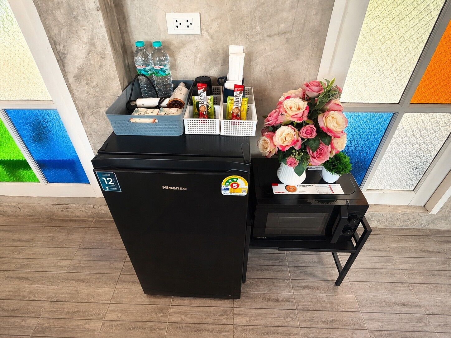 Uma geladeira compacta preta com o logo Hisense, uma pequena mesa preta com um monitor Toshiba, um vaso com flores rosa e brancas, e uma parede com textura.