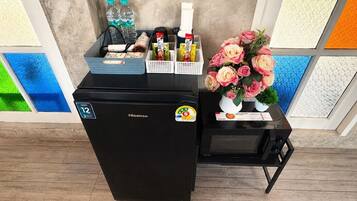 Uma geladeira compacta preta com o logo Hisense, uma pequena mesa preta com um monitor Toshiba, um vaso com flores rosa e brancas, e uma parede com textura.