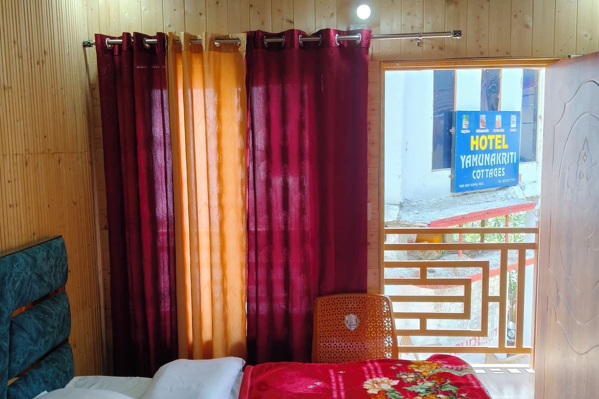 Quarto com painéis de madeira, uma cama, cortinas, uma cadeira e uma placa indicando Hotel Yamunakriti Cottages.