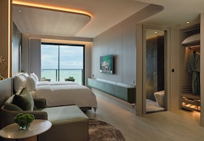 Quarto de hotel moderno com uma cama grande, área de estar com uma mesinha, televisão na parede e vista para o mar pela janela.