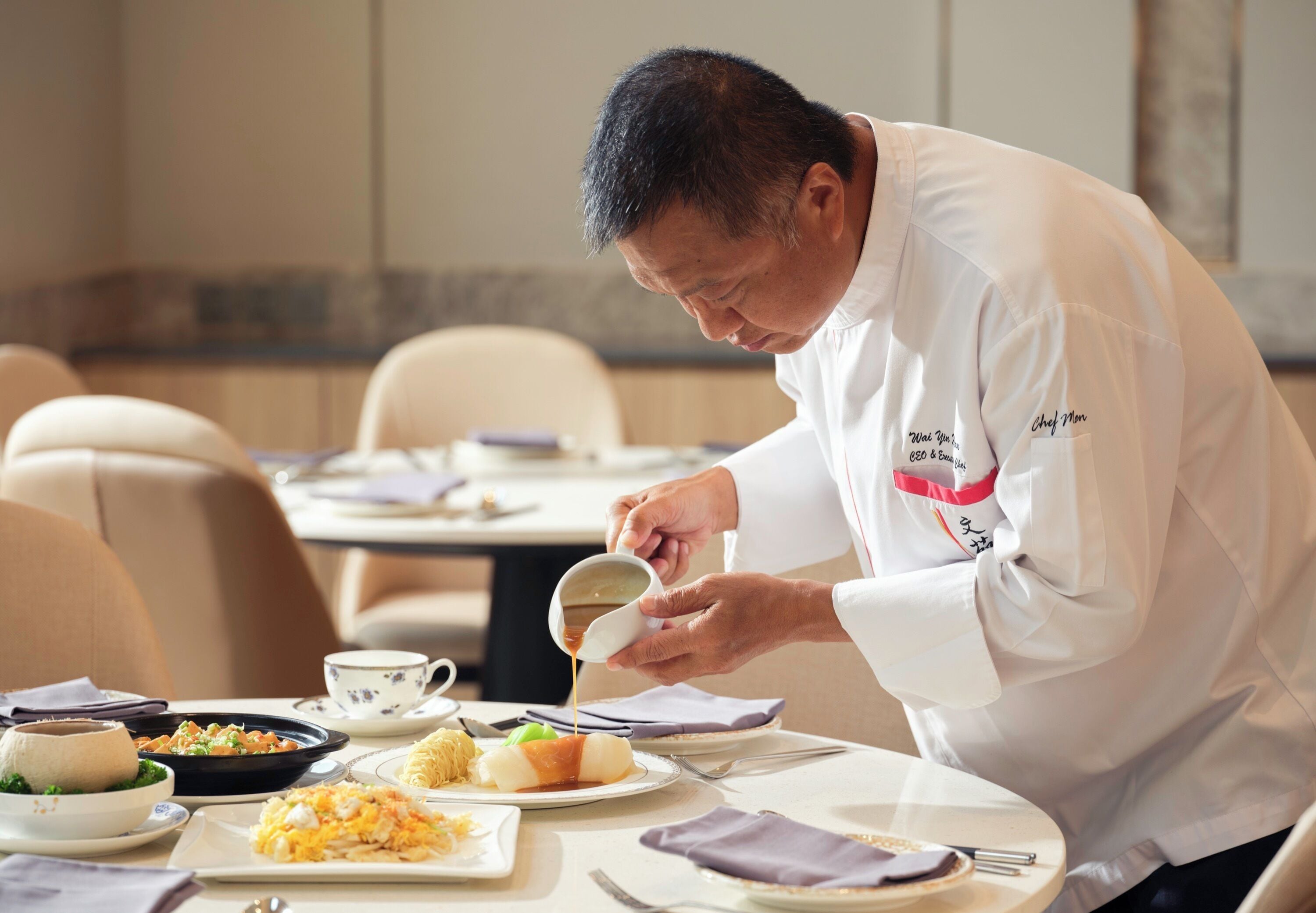 Um chef com uniforme branco despejando molho sobre um prato. Há pratos de comida, um bule e uma xícara na mesa.