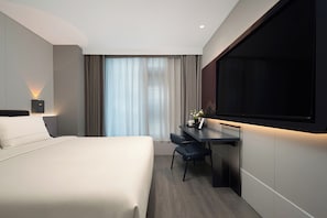 Quarto de hotel moderno com uma cama grande, uma escrivaninha com cadeira, uma TV de tela plana e uma janela com cortinas.