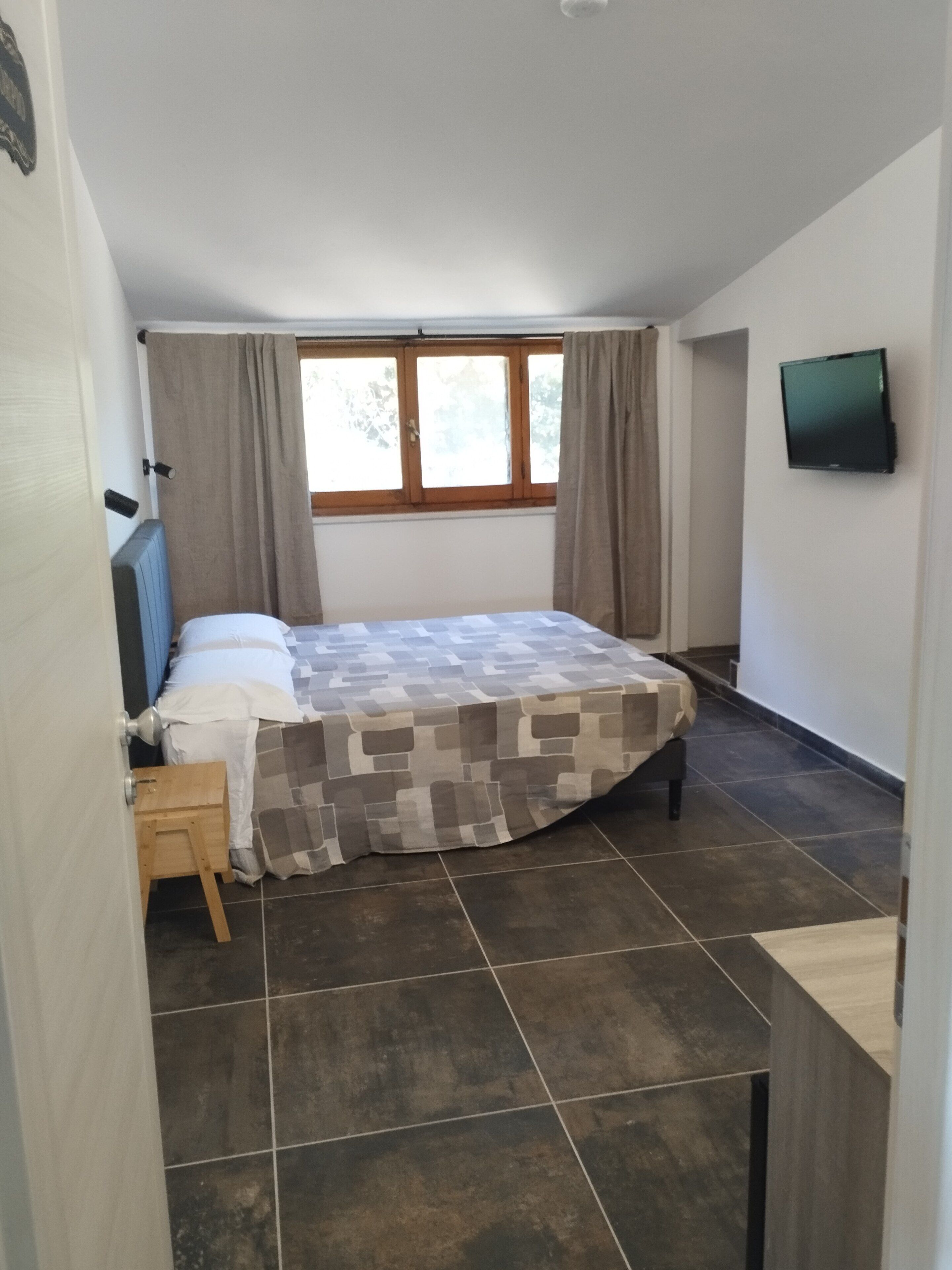 Quarto com cama, uma pequena mesa de madeira, televisão na parede e janela com cortinas.