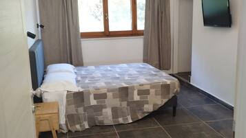 Quarto com cama, uma pequena mesa de madeira, televisão na parede e janela com cortinas.