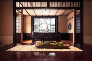 Um quarto japonês tradicional com piso de tatame, uma mesa de madeira baixa com uma tigela decorativa, um banco de madeira e uma porta deslizante com vista para árvores.