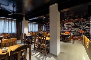 Interior de restaurante com mesas e cadeiras de madeira, parede preta e branca com ilustrações sobre café e grandes janelas com cortinas.