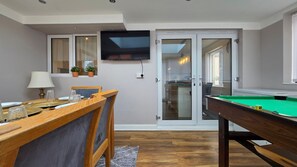 Quarto com mesa de bilhar, mesa de jantar com cadeiras, TV fixada na parede e porta de vidro deslizante que dá para uma cozinha.