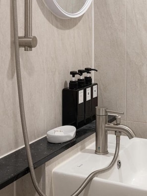 Casa de banho com lavatório branco, chuveiro manual e conjunto de produtos de higiene pessoal pretos.