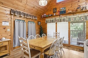 Interior de uma cabana de madeira com uma mesa de jantar, cadeiras, uma pequena escrivaninha e um pinheiro de Natal.