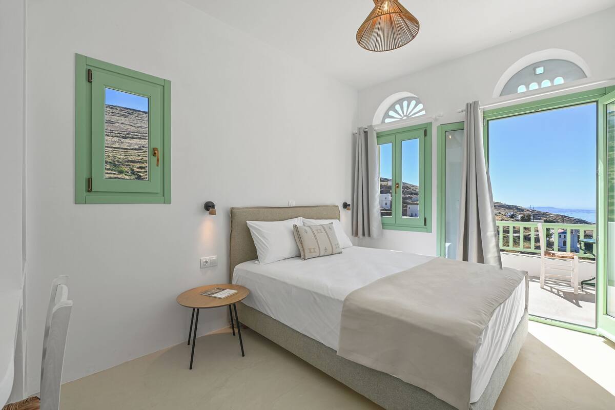 Quarto com cama, uma mesinha, luminária pendente e vista para o exterior.