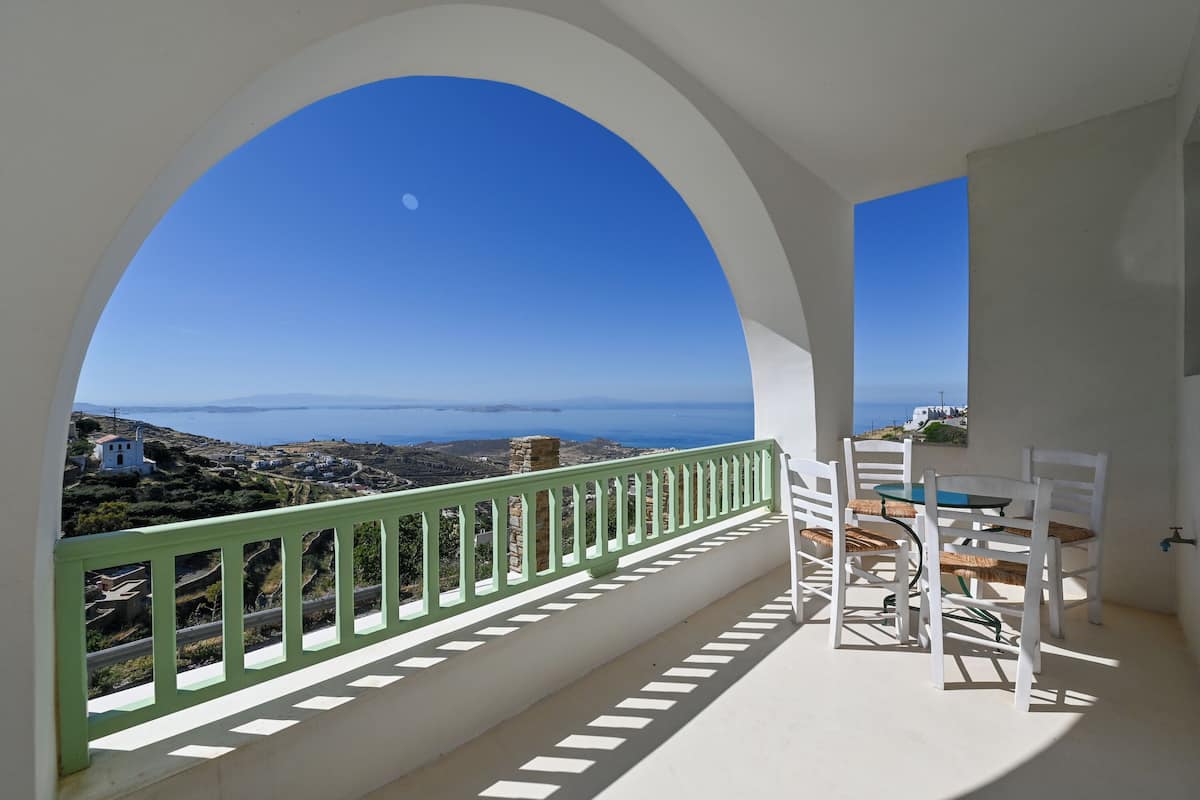 Uma varanda com vista para a paisagem costeira, uma mesa com cadeiras e um guarda-corpo verde.
