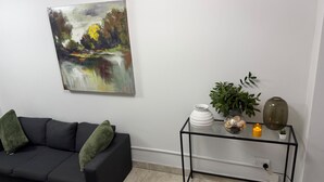Uma sala de estar com um sofá preto, uma mesa de centro de vidro, um quadro, um vaso de planta e um vaso.