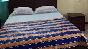 Uma cama com colcha estampada, mesa de cabeceira, ar-condicionado fixado na parede e janela com cortinas decorativas.