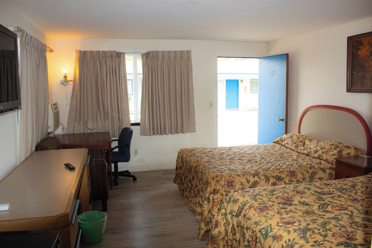 Quarto de hotel com cama, escrivaninha, cadeira, lixeira e porta que dá para o exterior.
