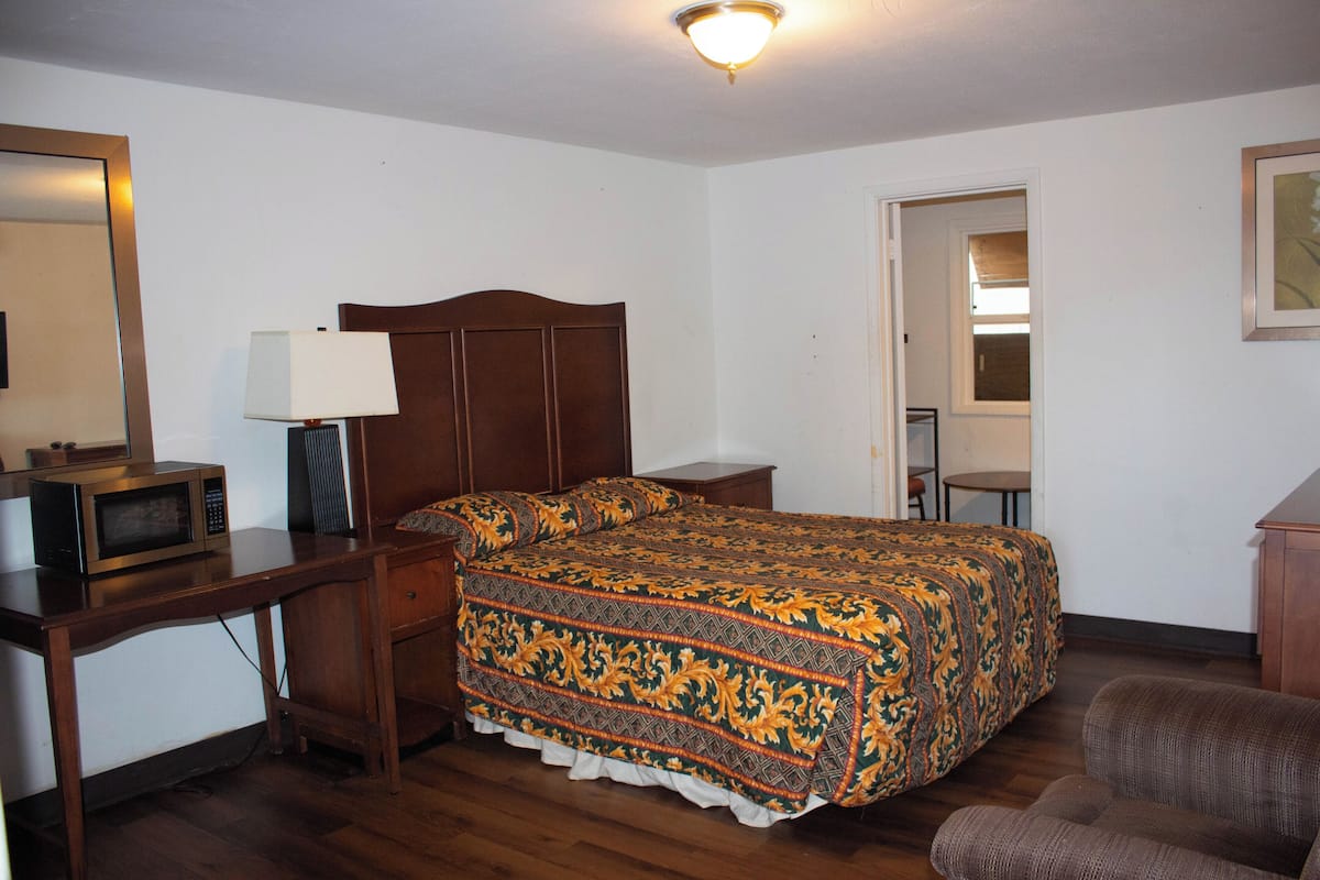 Quarto de hotel com cama de madeira, uma escrivaninha com micro-ondas, uma cadeira e uma porta que dá para outro cômodo.