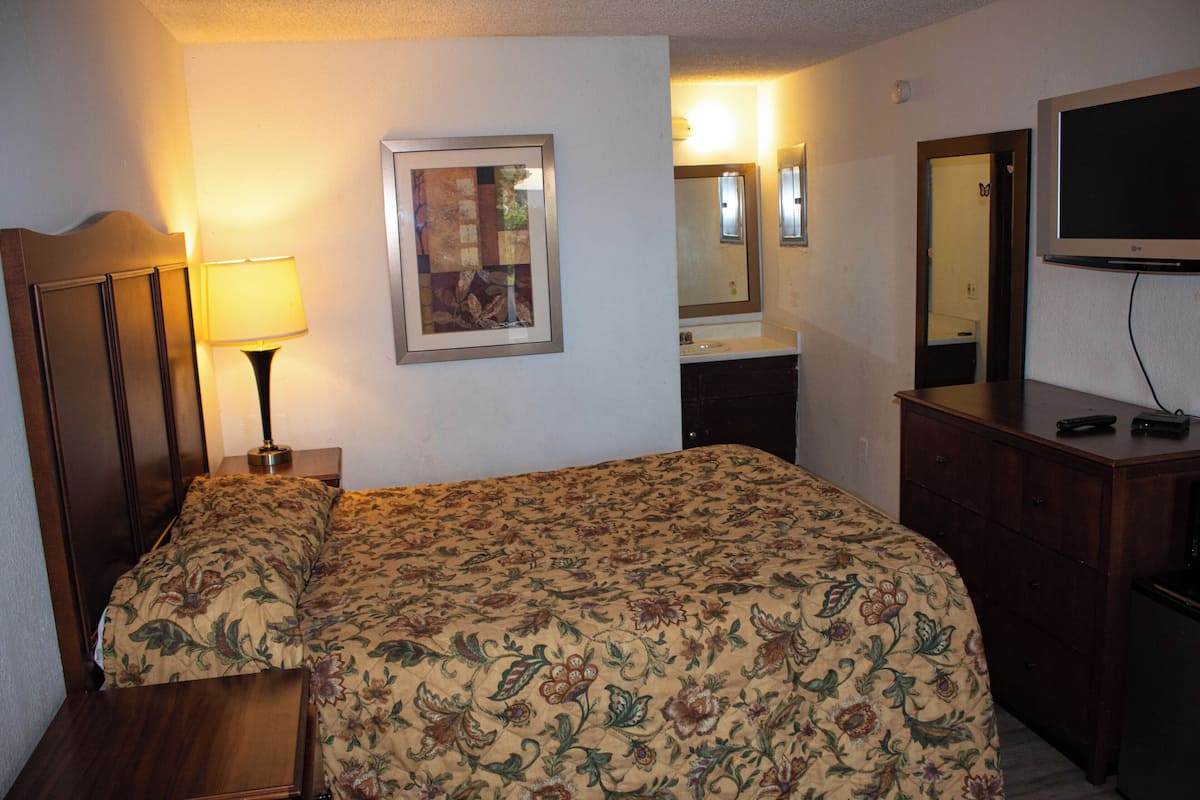 Quarto de hotel com cama, luminária, espelho, cômoda e televisão.