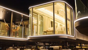 Uma estrutura de vidro moderna e iluminada, com interior visível, incluindo um sofá e uma mesa de jantar.