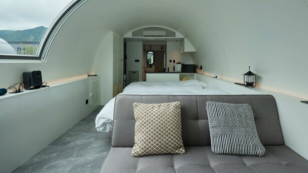 Quarto moderno e compacto com cama, duas almofadas estampadas, janela e vista para uma montanha.