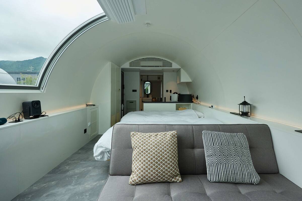 Quarto moderno e compacto com cama, duas almofadas estampadas, janela e vista para uma montanha.