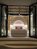 Quarto moderno com cama centralizada sob uma grande janela em arco, um ventilador de teto e vista para um ambiente tradicional com telhado de palha.