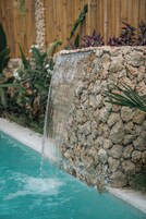 Uma cascata de pedra deságua em uma piscina, cercada por plantas e um cercado de bambu.