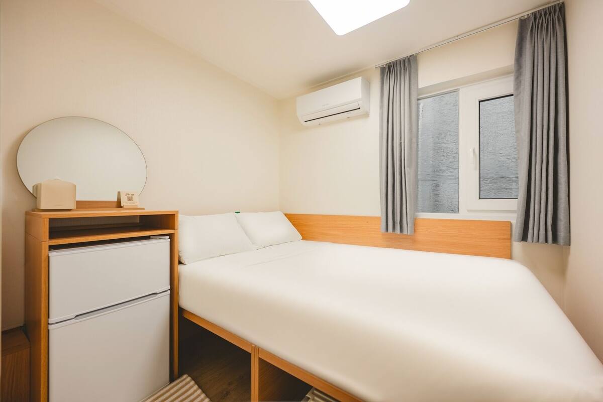 Quarto de hotel com uma cama, uma pequena escrivaninha com espelho, um refrigerador e um aparelho de ar condicionado.