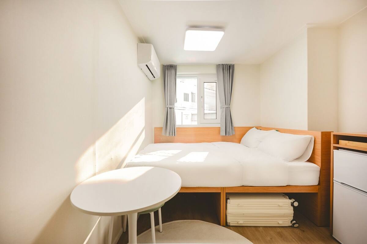 Quarto de hotel pequeno com uma cama, uma mesinha, um radiador e uma janela com cortinas.