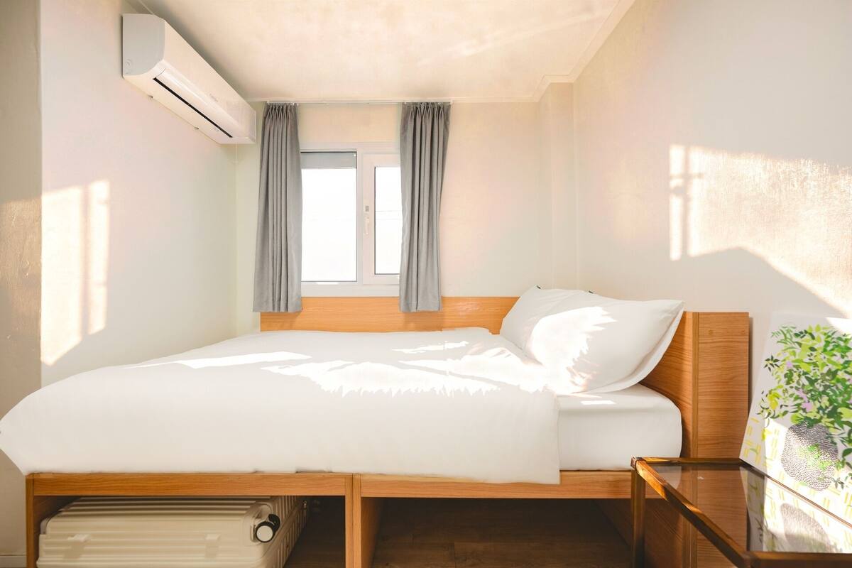 Quarto de hotel com cama, unidade de ar condicionado, janela com cortinas e mesa de cabeceira com uma planta.