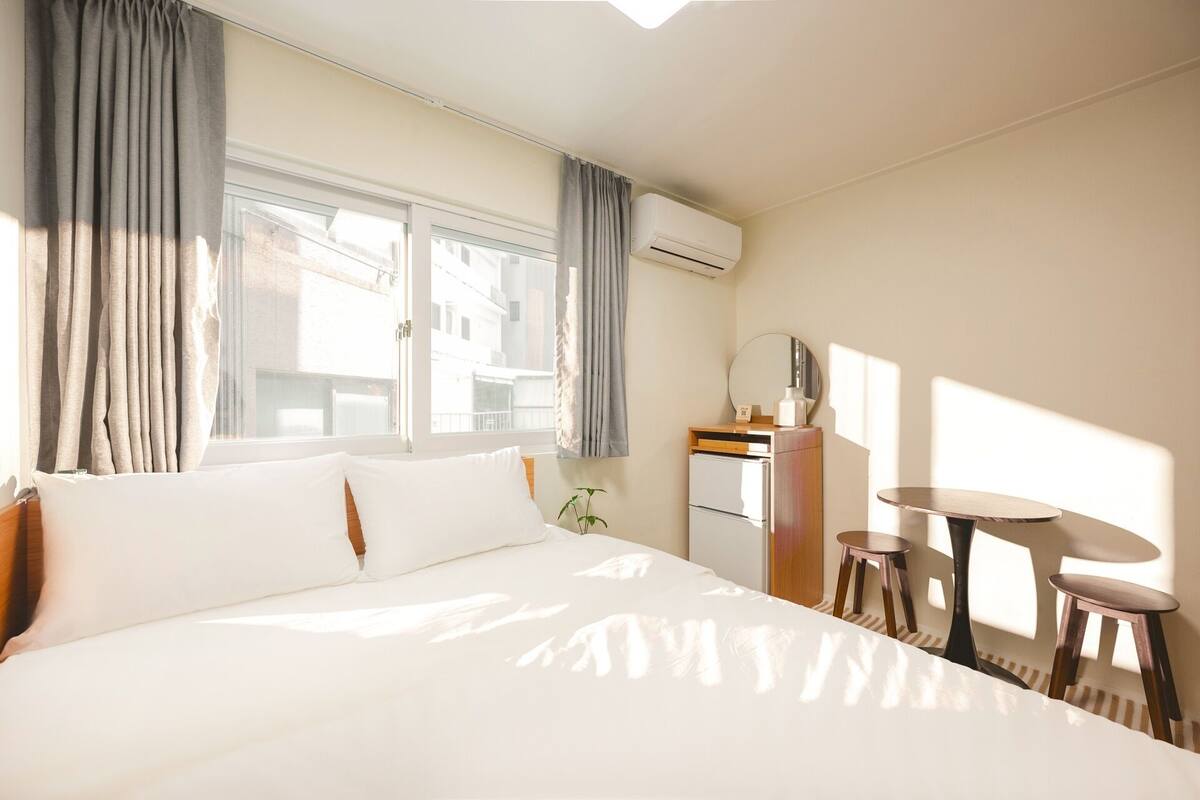 Quarto de hotel com cama, uma pequena mesa, uma cadeira, ar condicionado, espelho e janela com cortinas.