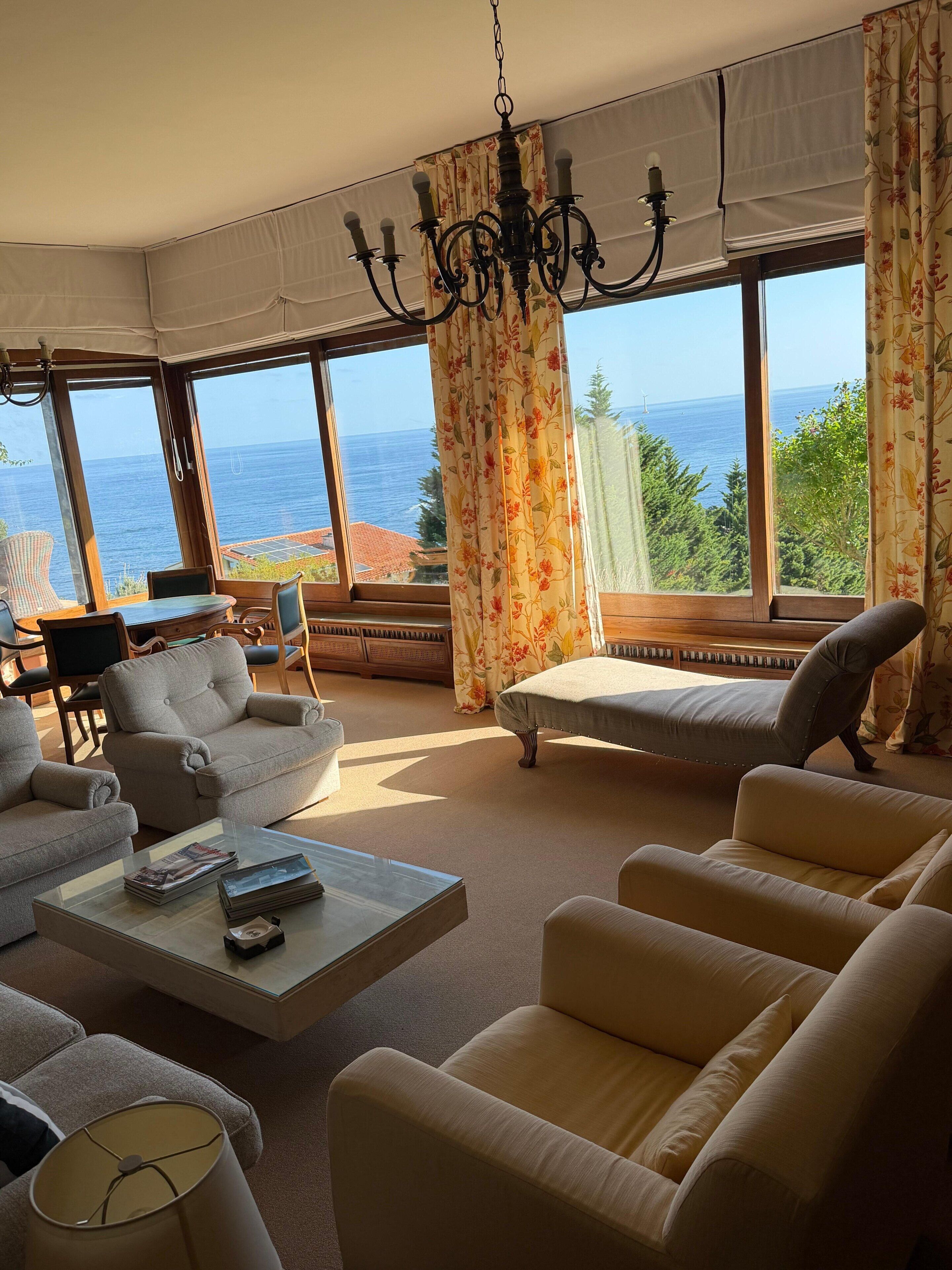 Uma sala de estar ampla com uma grande janela com vista para o mar, um candelabro e um conjunto de sofás.