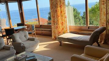 Uma sala de estar ampla com uma grande janela com vista para o mar, um candelabro e um conjunto de sofás.