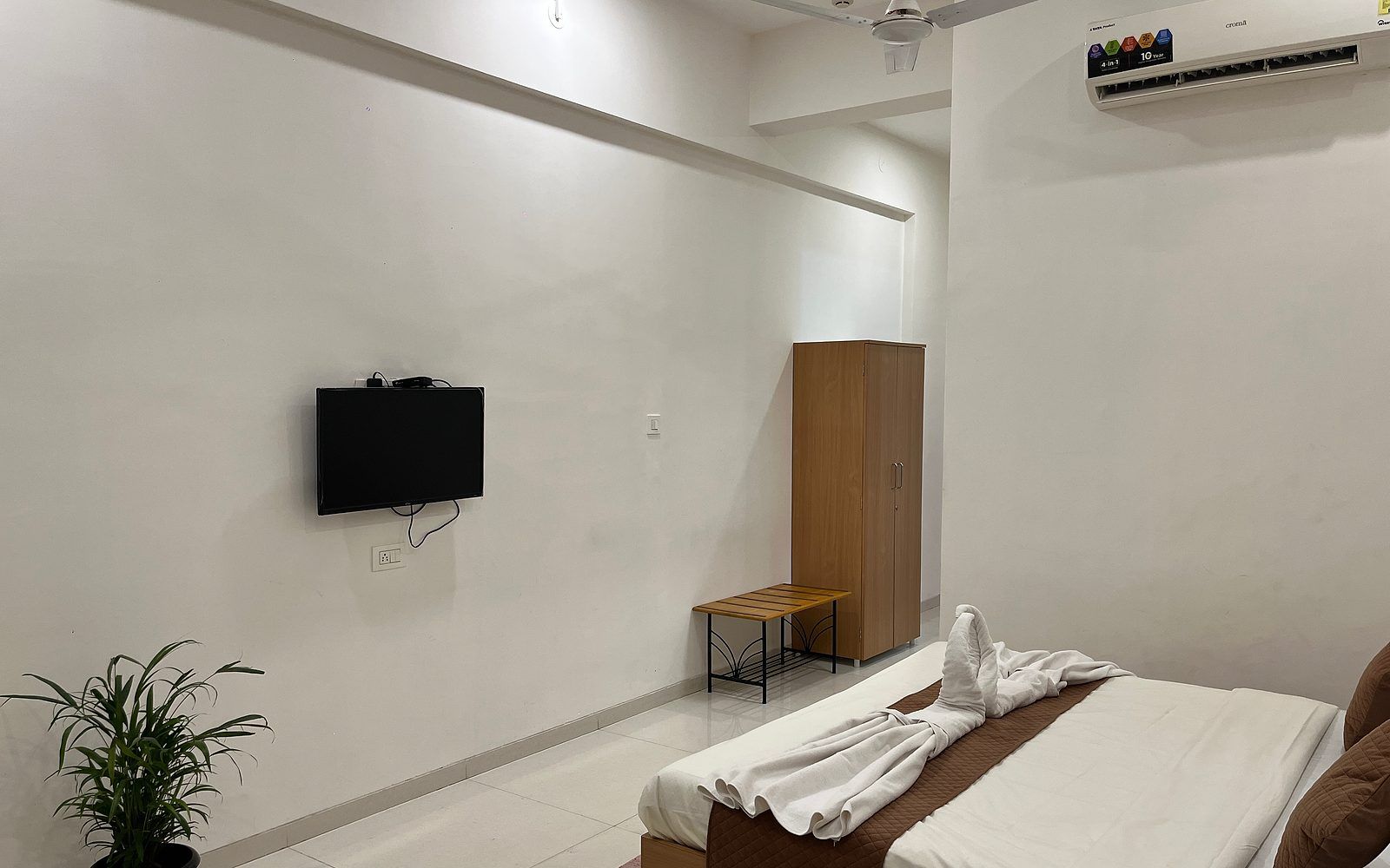 Quarto de hotel com cama, televisão na parede, guarda-roupa, mesa pequena e planta em vaso.