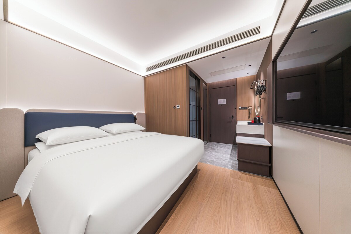 Quarto de hotel moderno com uma cama grande, banheiro com espelho e uma pequena cozinha.