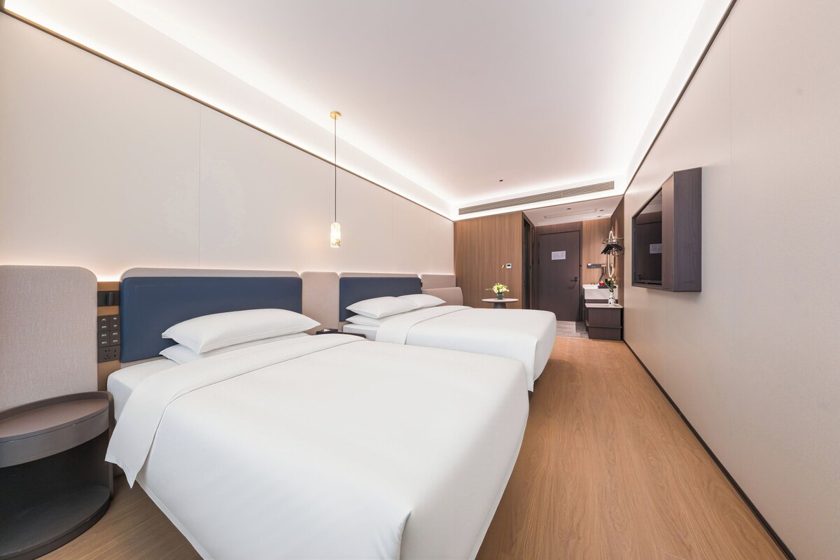 Quarto de hotel com três camas, uma mesinha, televisão presa à parede e porta para o banheiro.