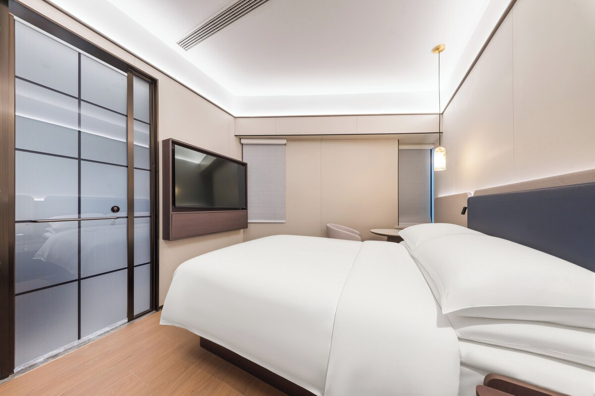 Quarto de hotel moderno com uma cama grande, televisão de tela plana, porta deslizante e uma pequena mesa.