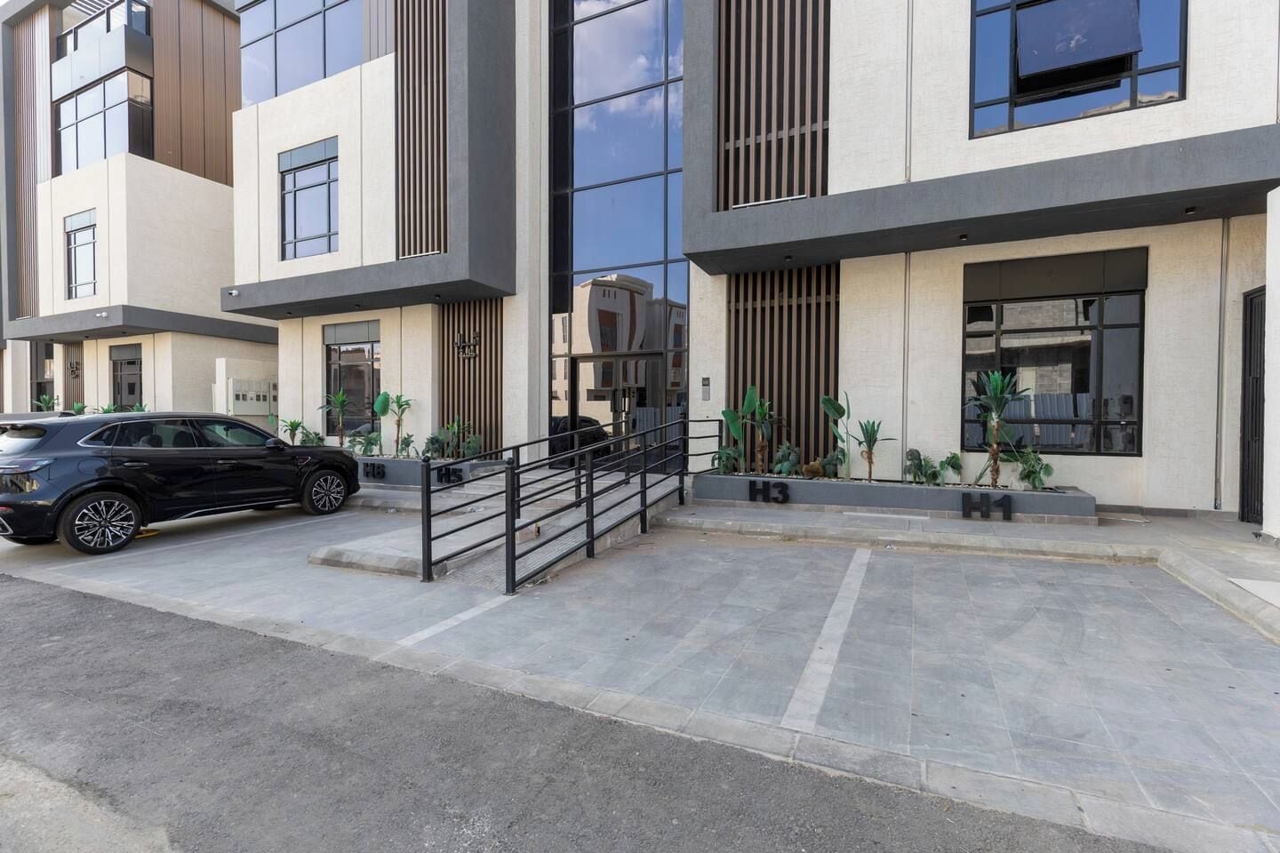 Um edifício moderno com um carro preto estacionado na frente, uma escadaria que leva à entrada e uma pequena área de jardim com plantas.