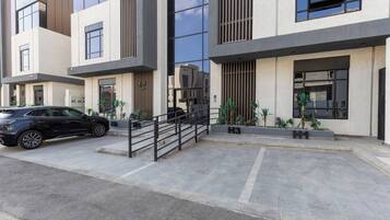 Um edifício moderno com um carro preto estacionado na frente, uma escadaria que leva à entrada e uma pequena área de jardim com plantas.