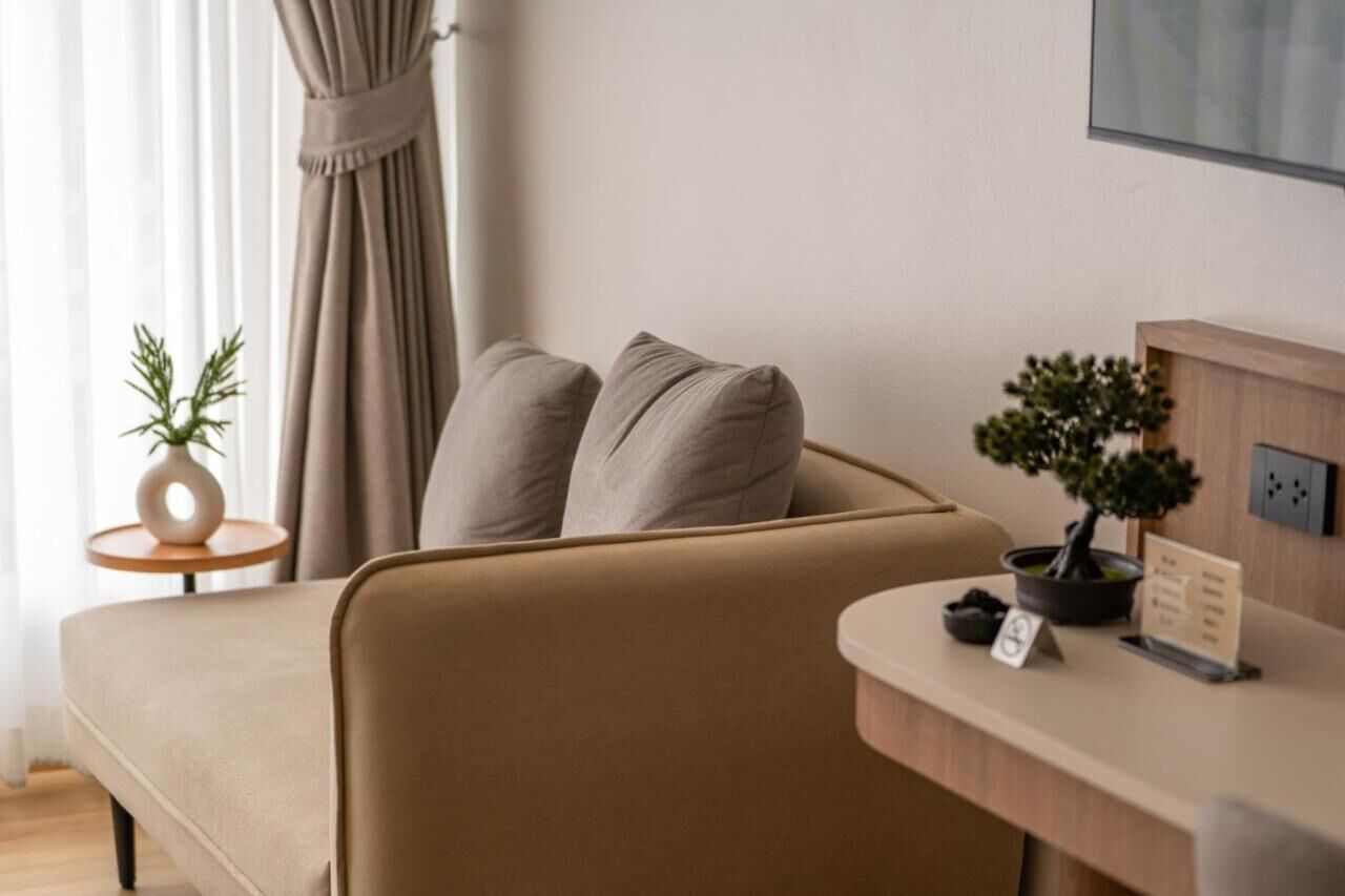 Quarto de hotel moderno com sofá, uma pequena mesa redonda com uma planta, uma planta em vaso sobre uma escrivaninha e uma TV fixada na parede.
