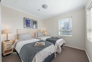 Quarto com duas camas, mesa de cabeceira com abajur, janela com persianas e quadros na parede.